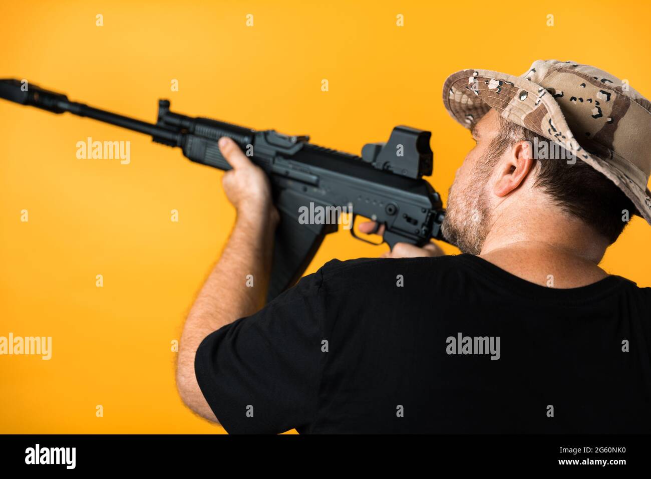 Middle aged man with rifle in black t-shirt and hat against yellow ...