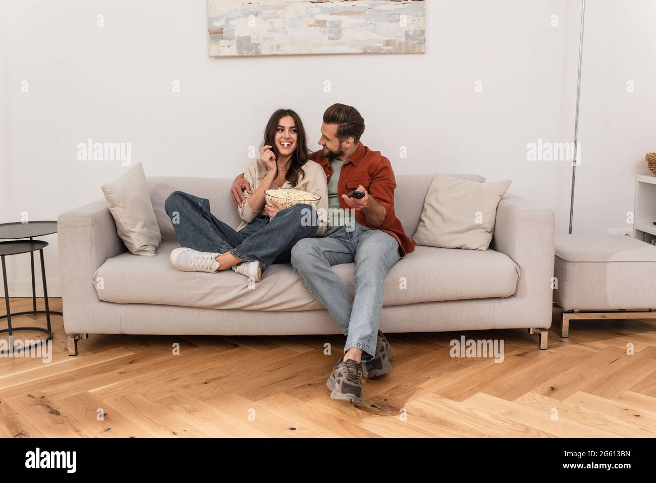 Smiling man with remote controller looking at girlfriend eating popcorn ...