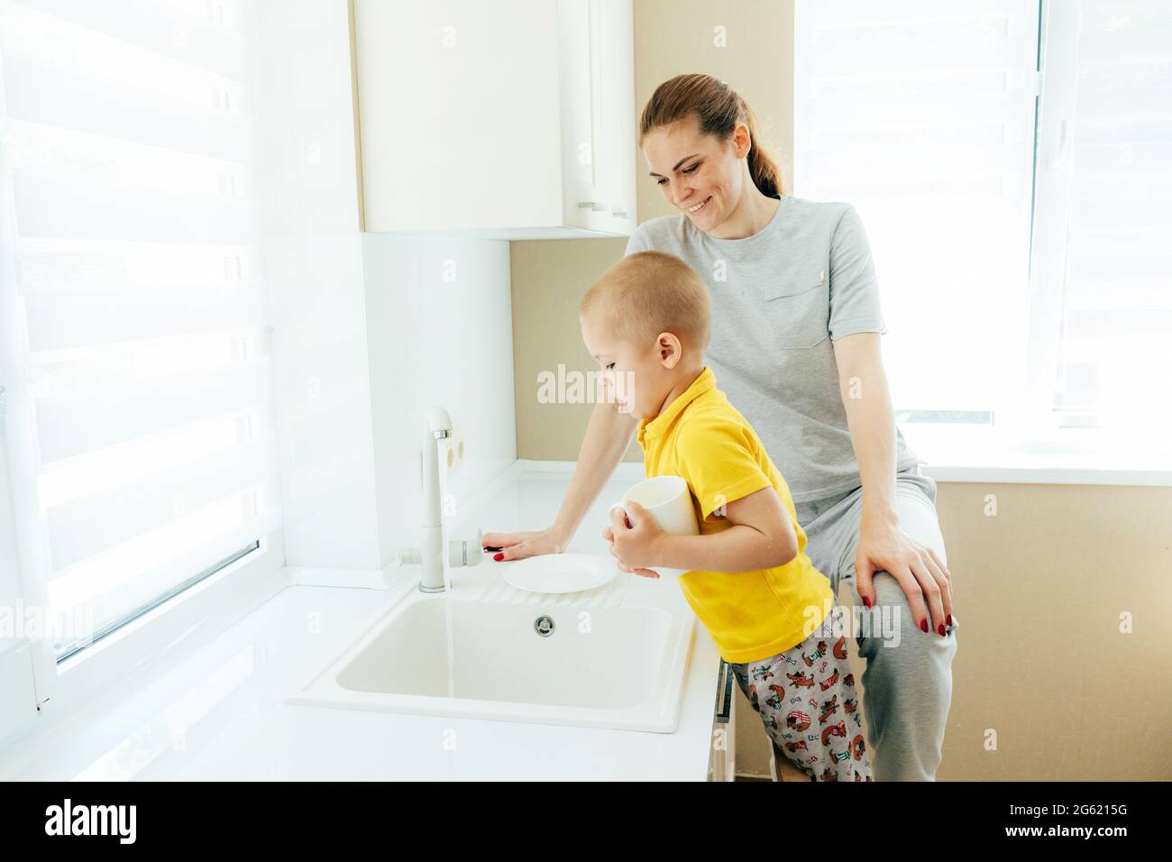 Happy mom teaches little son to wash dishes in the sink. Toddler and his mother washing dishes ...