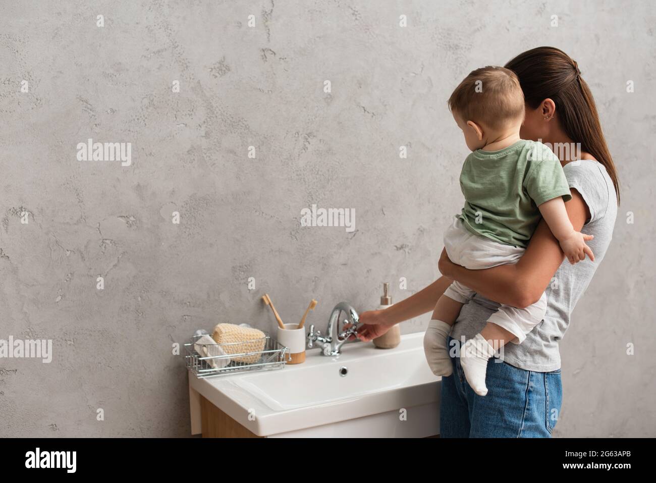 Mother opening tap of faucet while holding toddler kid in bathroom ...