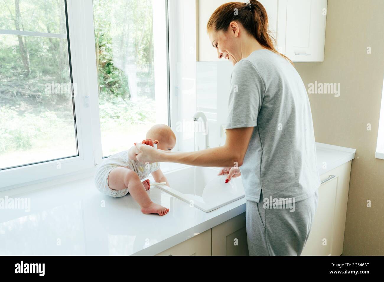 A young active mommy with a baby wash the dishes in the kitchen. While ...