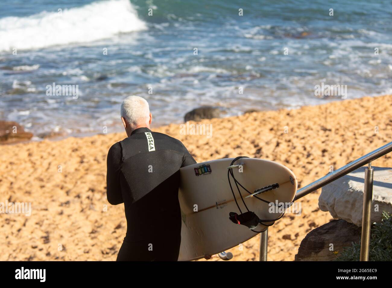 Middle aged senior male surfer wearing wetsuit carries surfboard to the ...