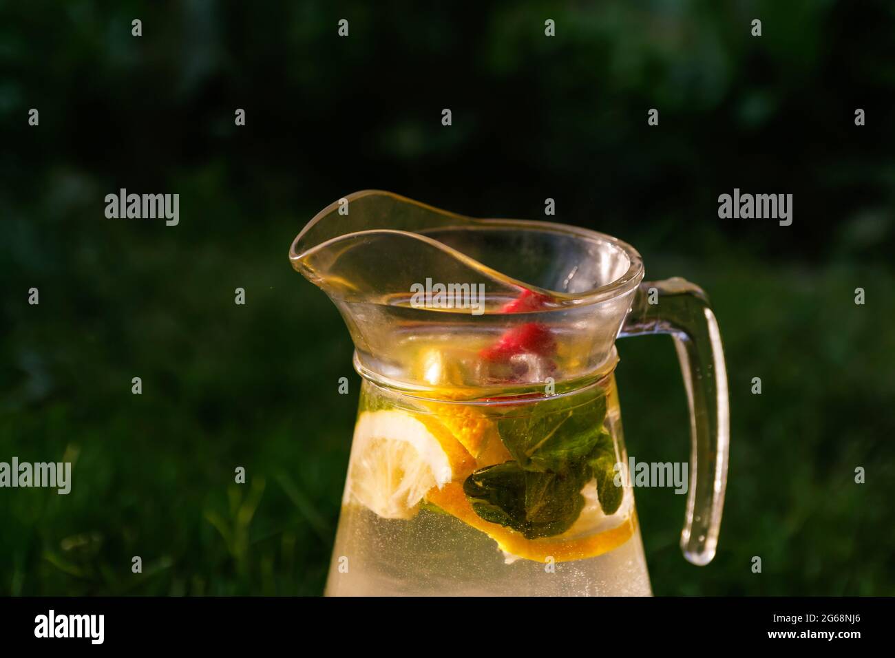 Defocus glass jug of lemonade with slice lemon, strawberry and mint on natural deep green ...