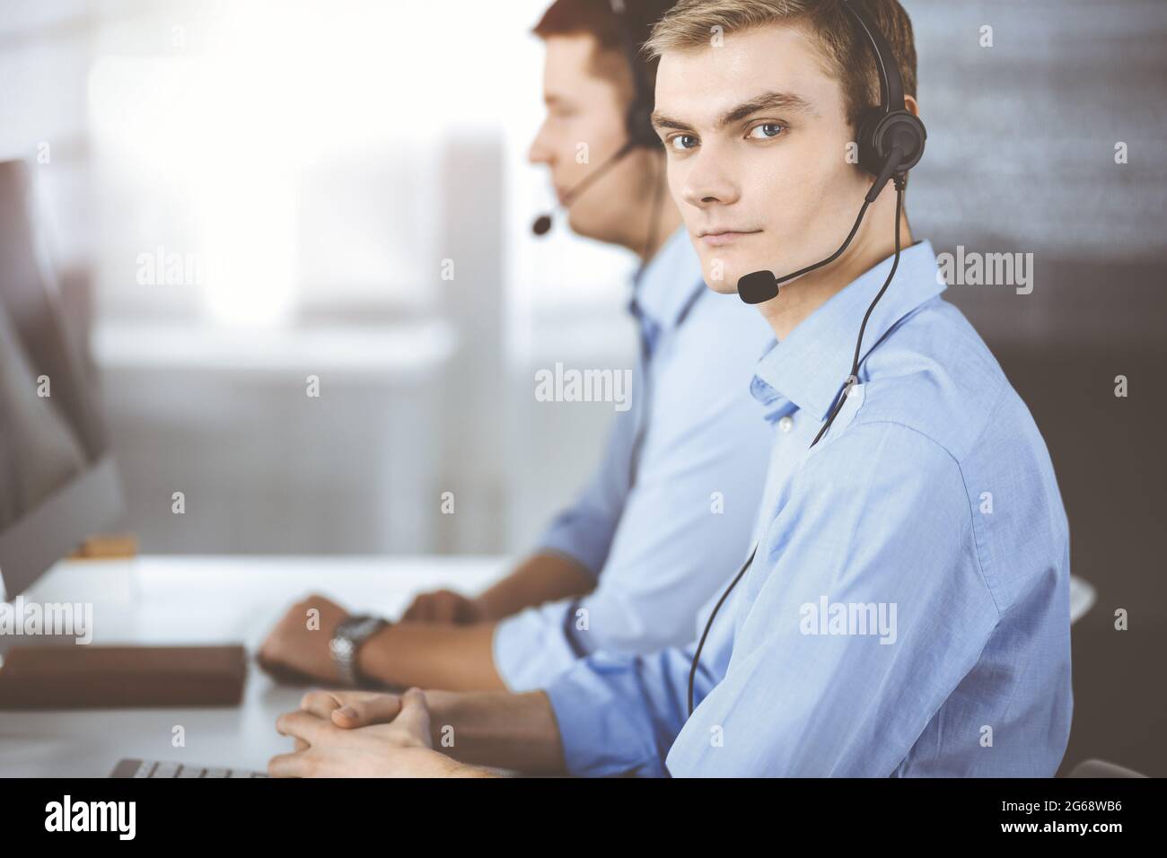 Two young men in headset, sitting at the desk in a sunny modern office ...