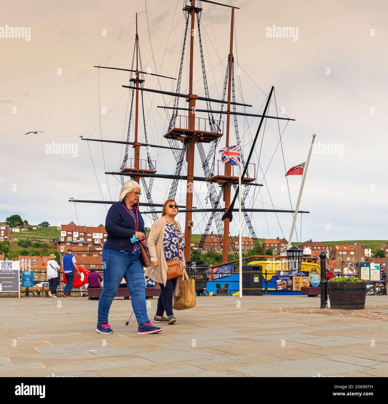 Two ladies walk along a quayside in front of a replica of a tall ship ...