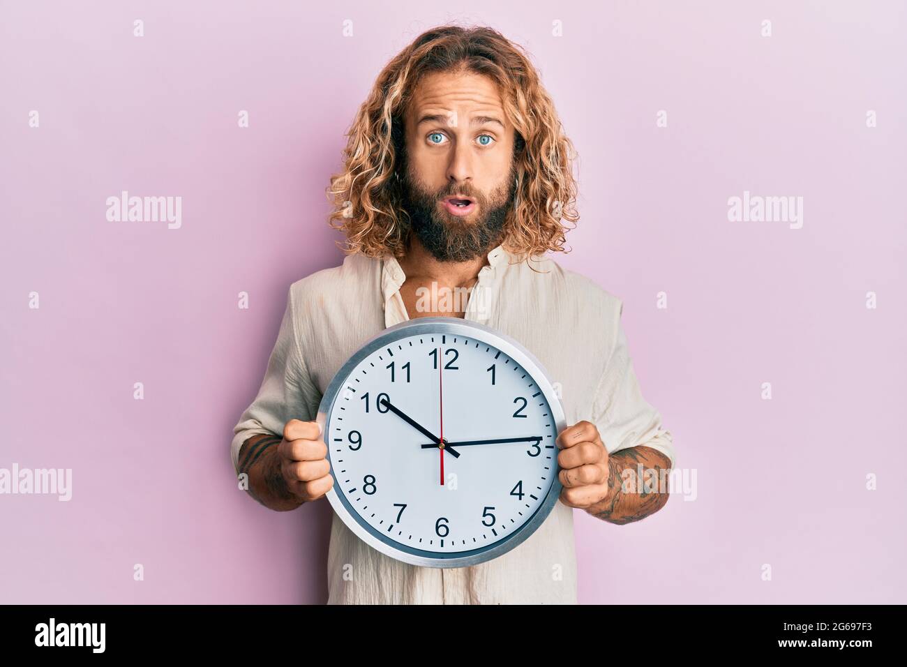 Handsome man with beard and long hair holding big clock in shock face ...