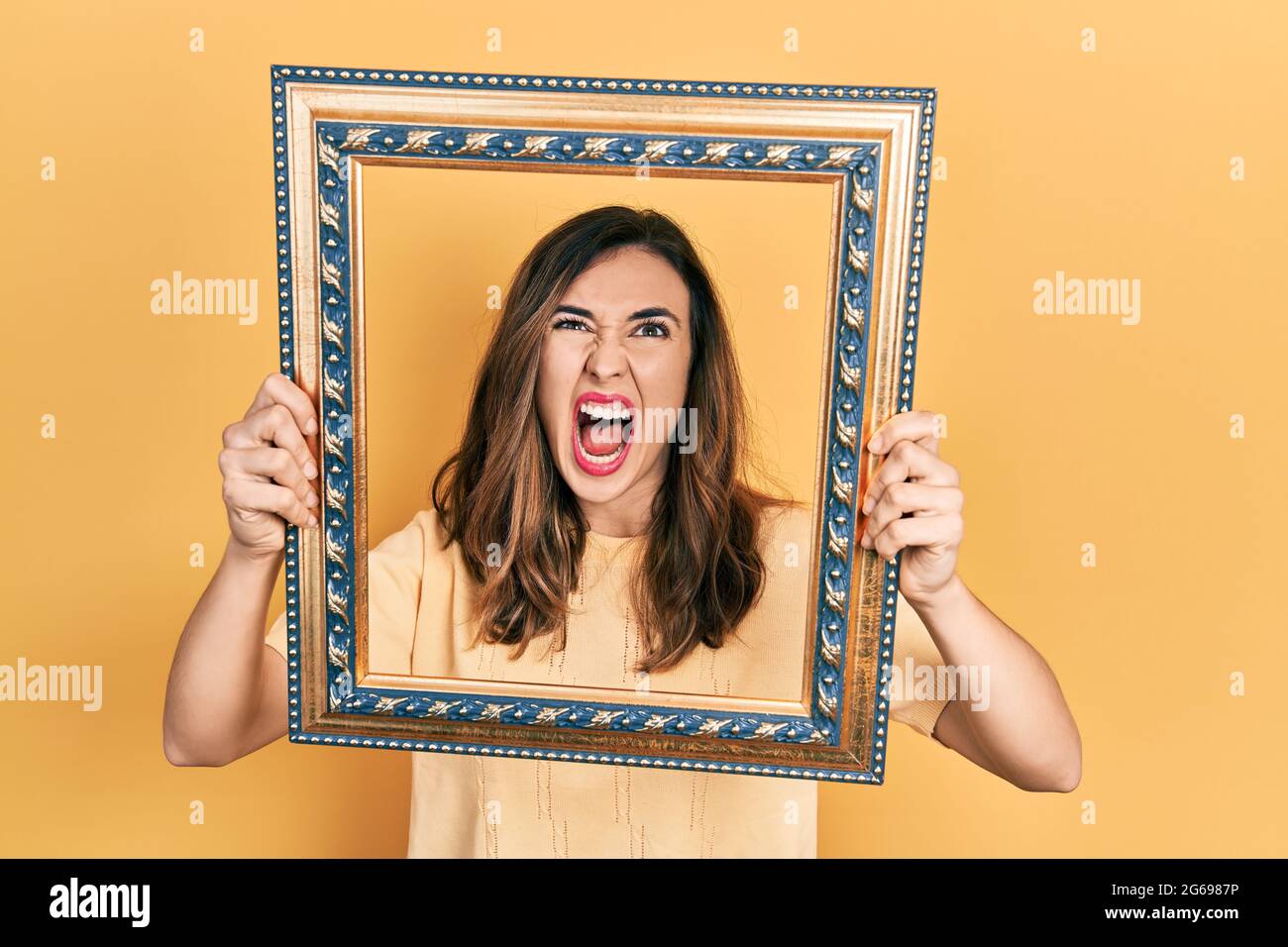 Young hispanic girl holding empty frame angry and mad screaming ...