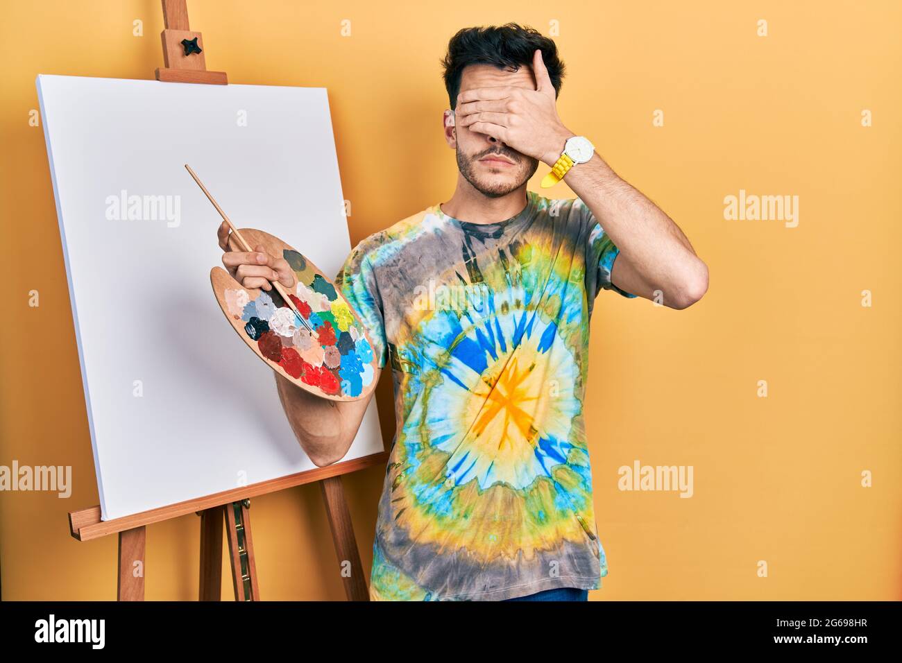 Young hispanic man standing drawing with palette by painter easel stand ...