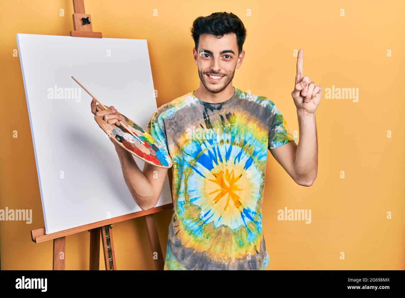 Young hispanic man standing drawing with palette by painter easel stand ...