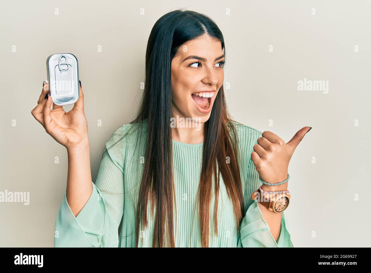Young hispanic woman holding canned food pointing thumb up to the side ...
