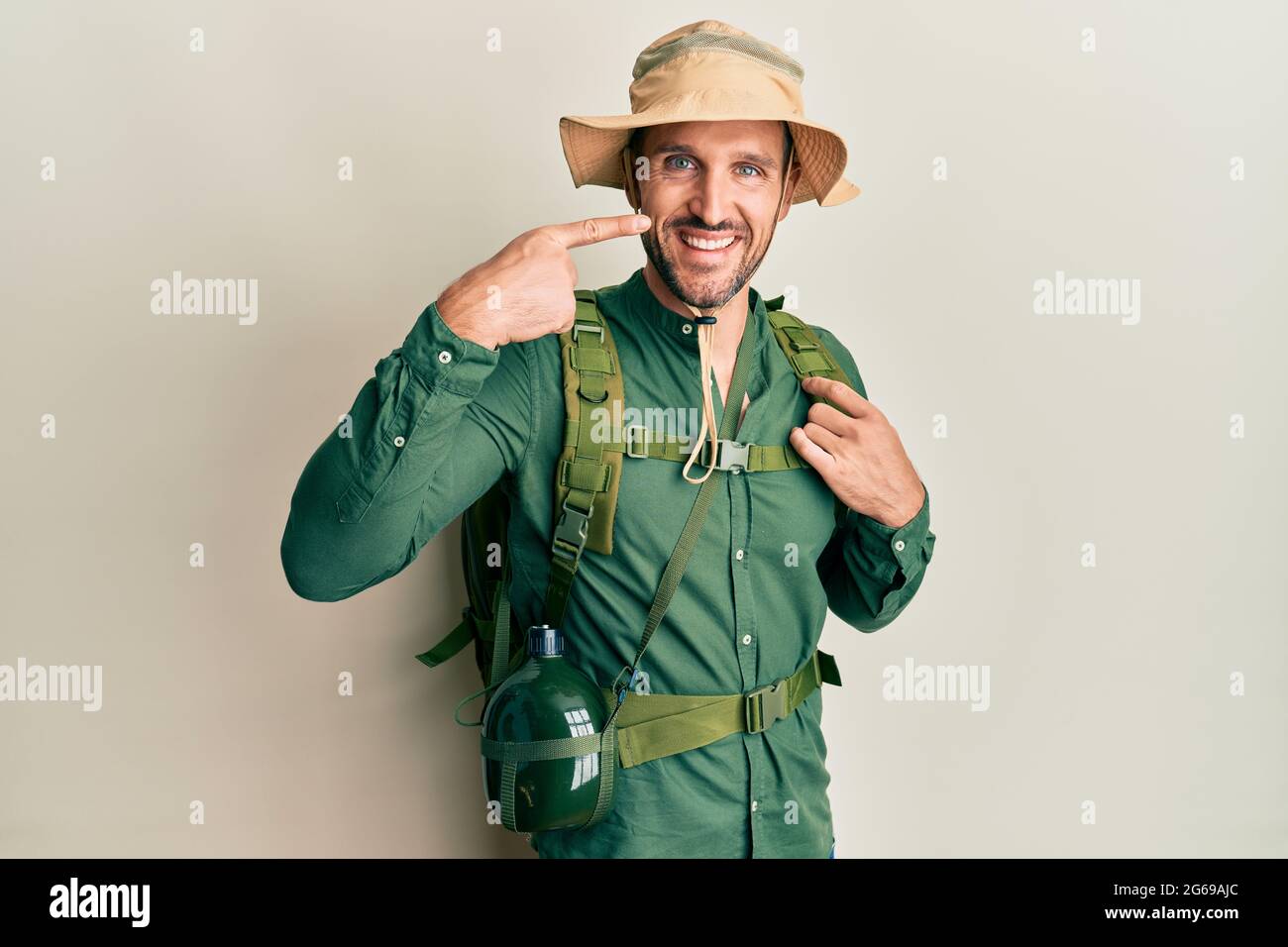 Handsome man with beard wearing explorer hat and backpack pointing with ...