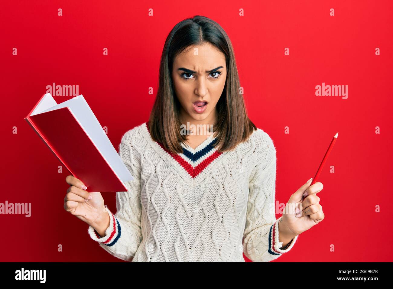 Young brunette girl holding book and pencil in shock face, looking ...