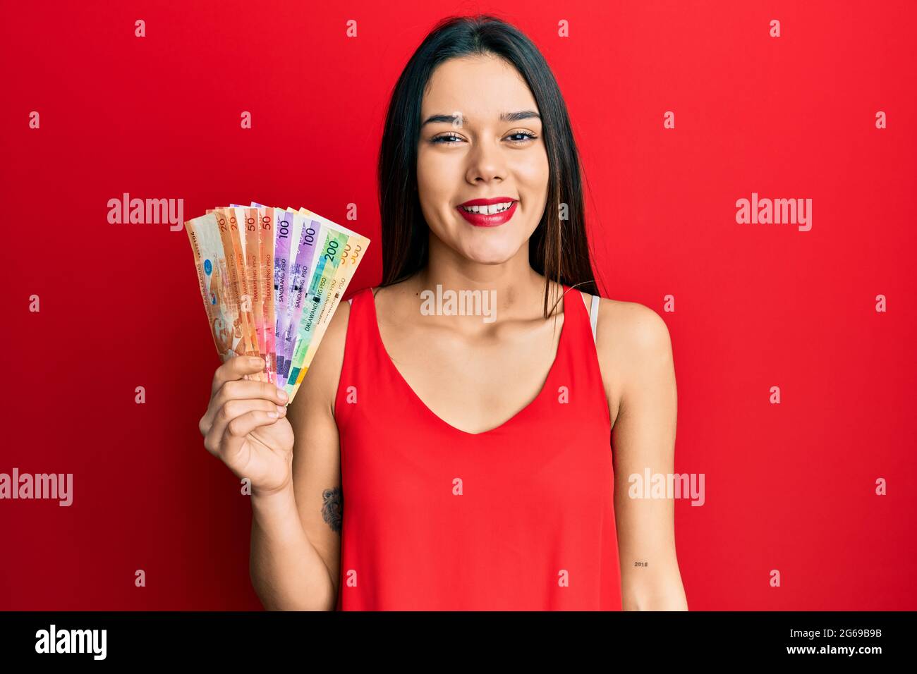 Young hispanic girl holding philippine peso banknotes looking positive ...