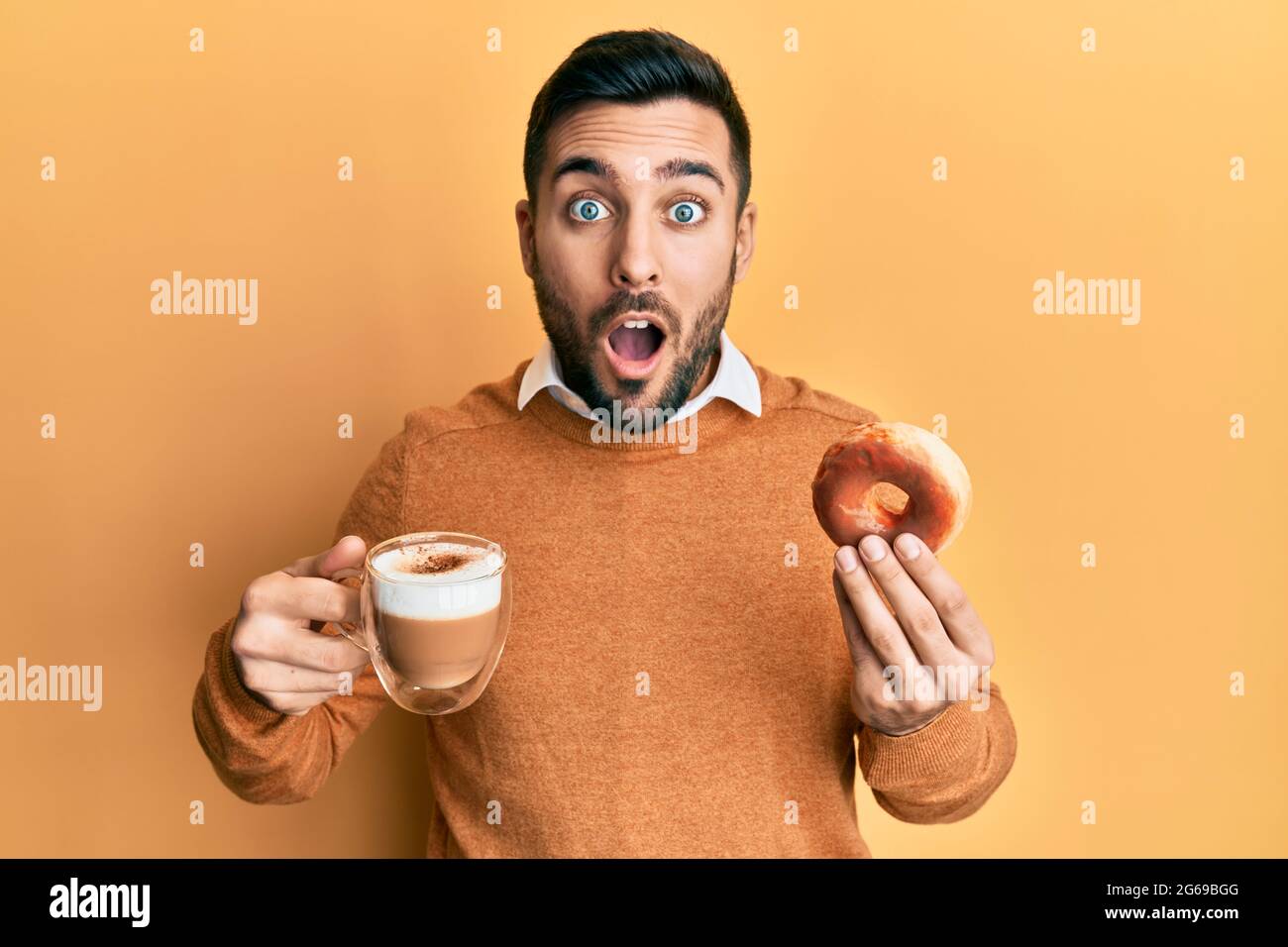 Young hispanic man eating doughnut and drinking coffee afraid and ...