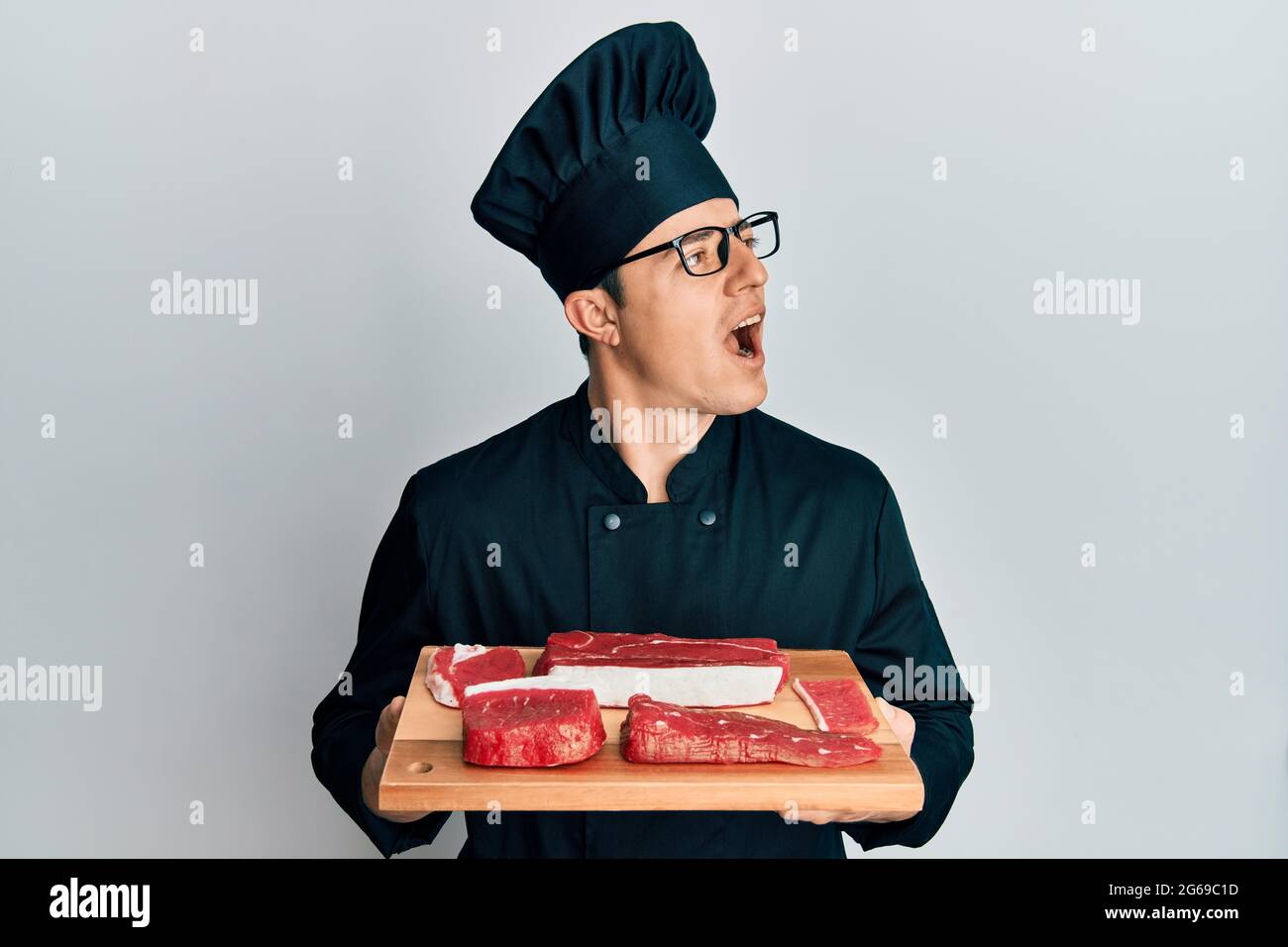 Handsome young man wearing chef uniform holding board with raw meat ...