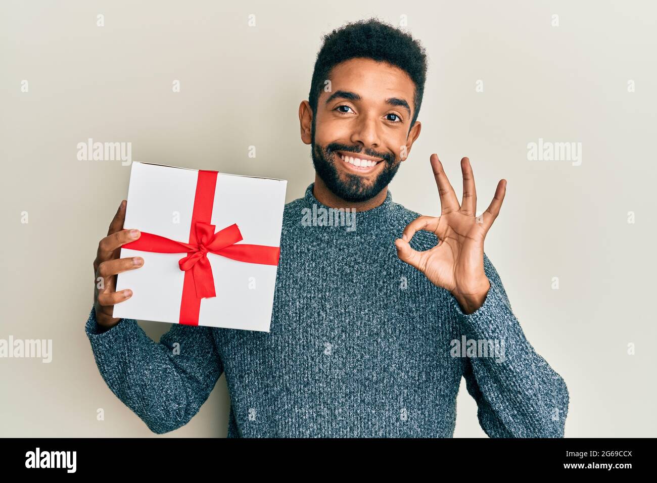 Handsome hispanic man with beard holding gift doing ok sign with ...