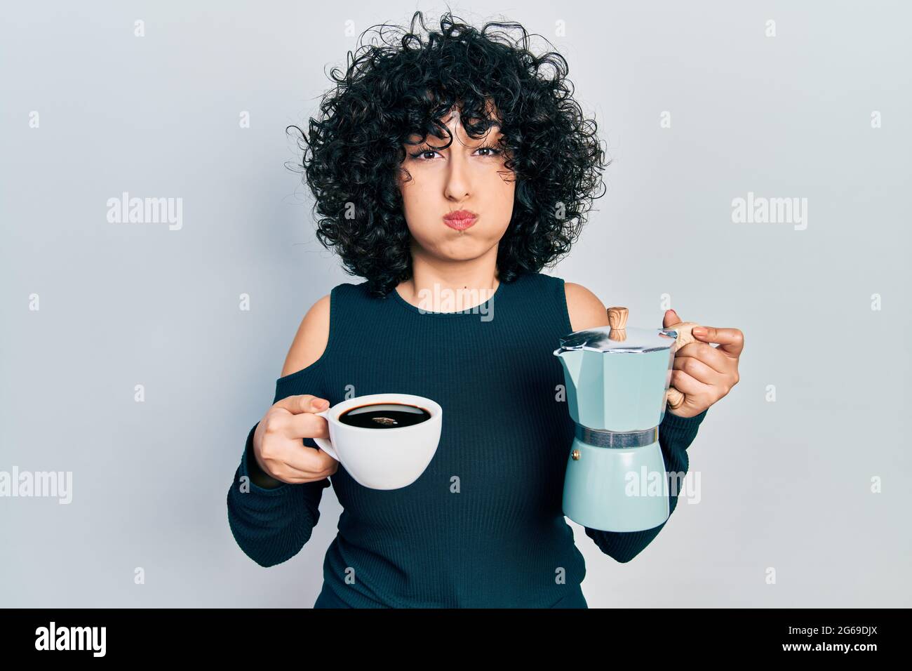 Young middle east woman holding italian coffee maker puffing cheeks ...