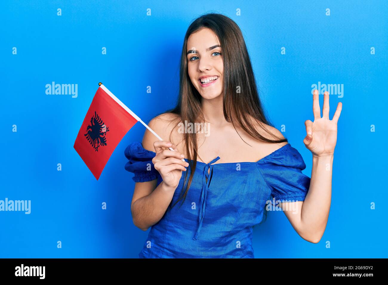 Young brunette teenager holding albania flag doing ok sign with fingers ...