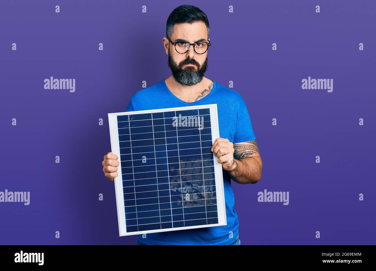 Hispanic man with beard holding photovoltaic solar panel depressed and ...