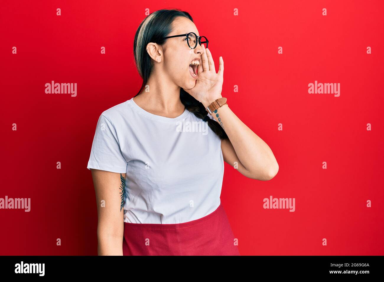 Young hispanic woman wearing professional waitress apron shouting and ...