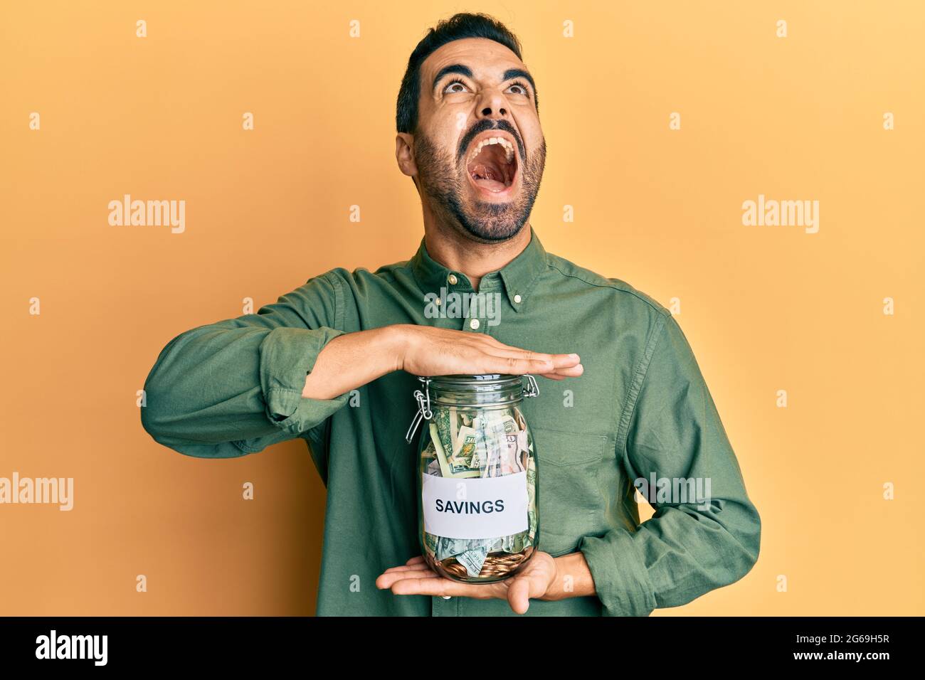 Young hispanic man holding jar with savings angry and mad screaming ...