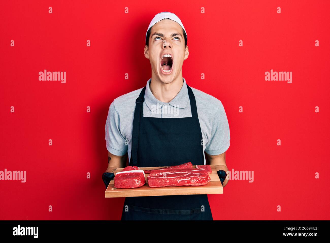 Young hispanic man holding board with raw meat angry and mad screaming ...