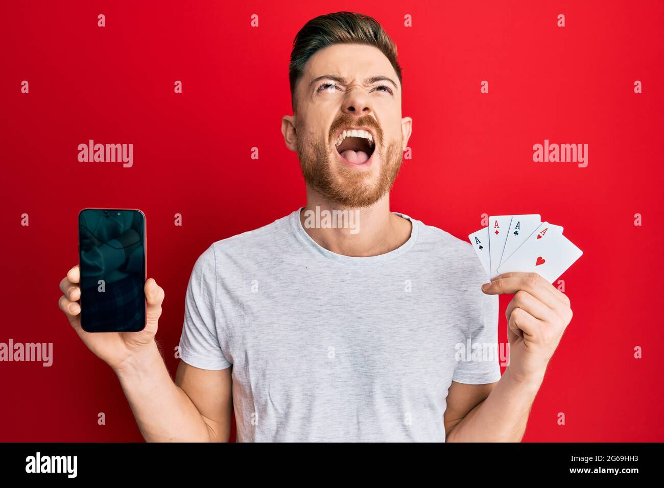 Young redhead man holding smartphone and casino card angry and mad ...