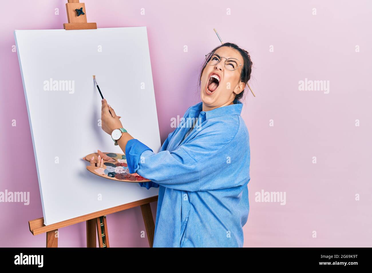 Young hispanic woman standing drawing with palette by painter easel ...