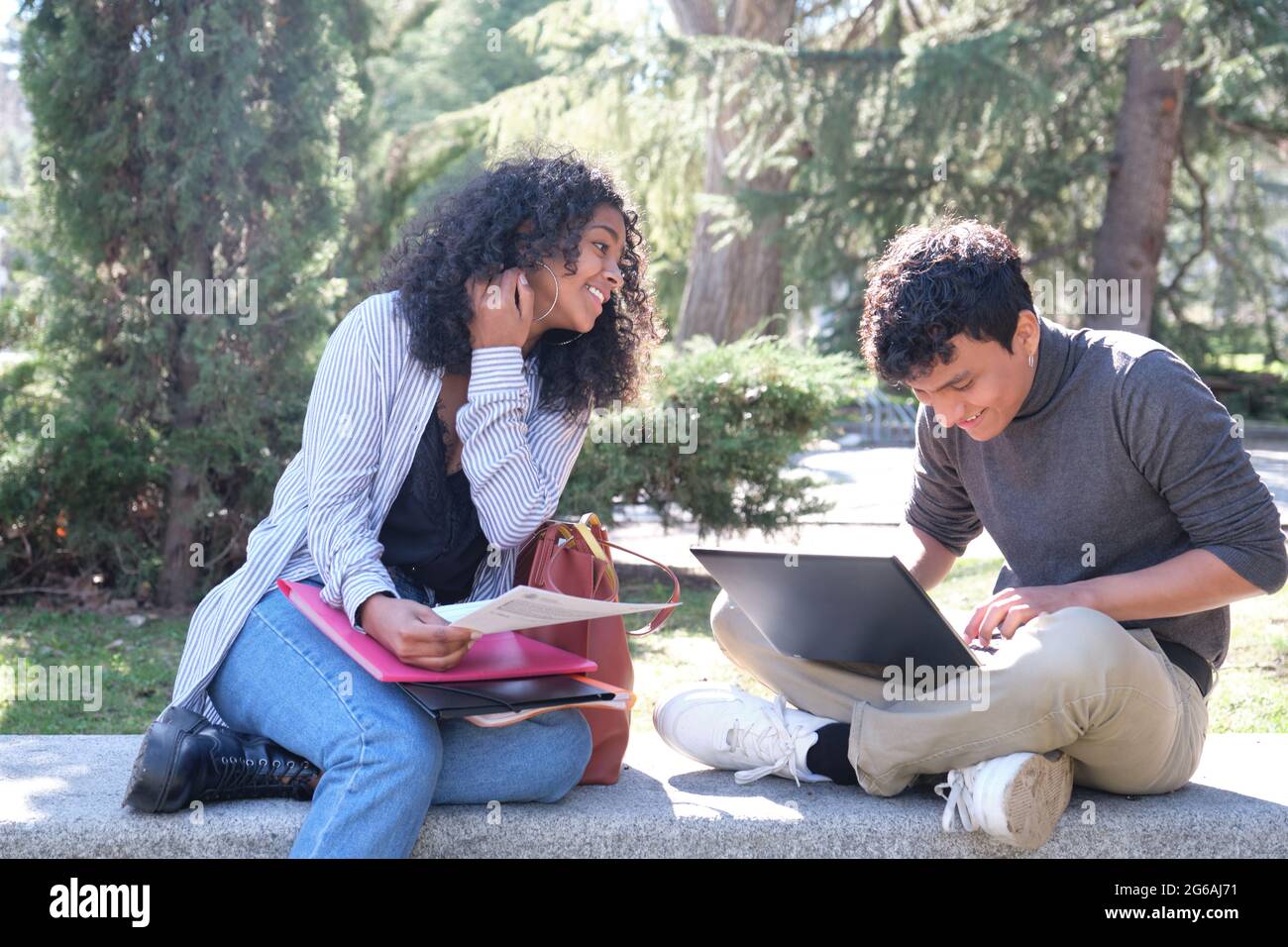 Two latin students smiling studying together sitting on a bench ...