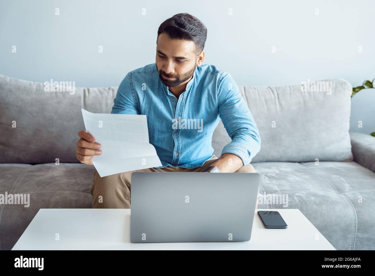 Focused serious indian man reading paper letter sitting front of laptop ...