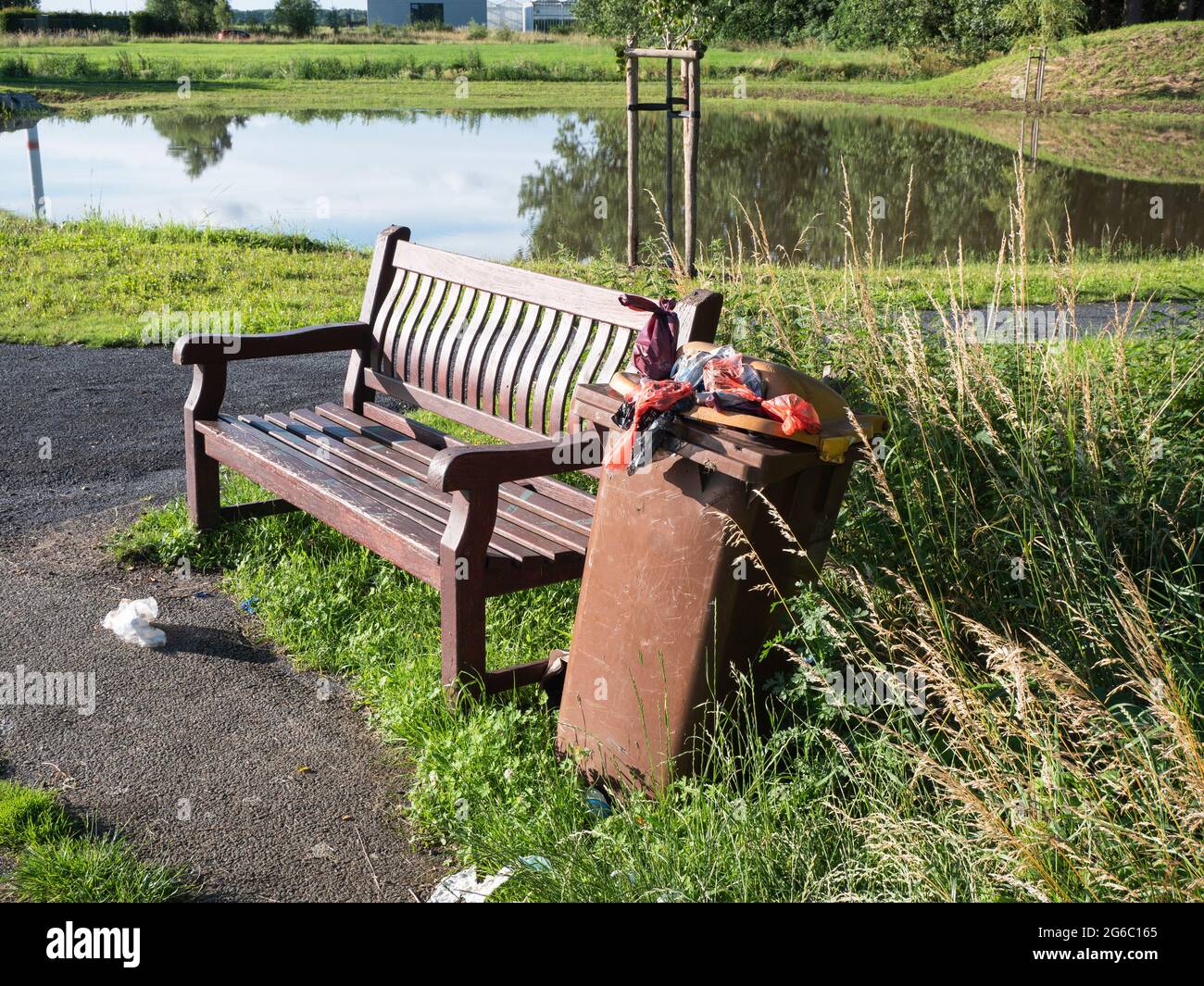 Wooden bench in a park with a pond and an overflowing trash can next to ...
