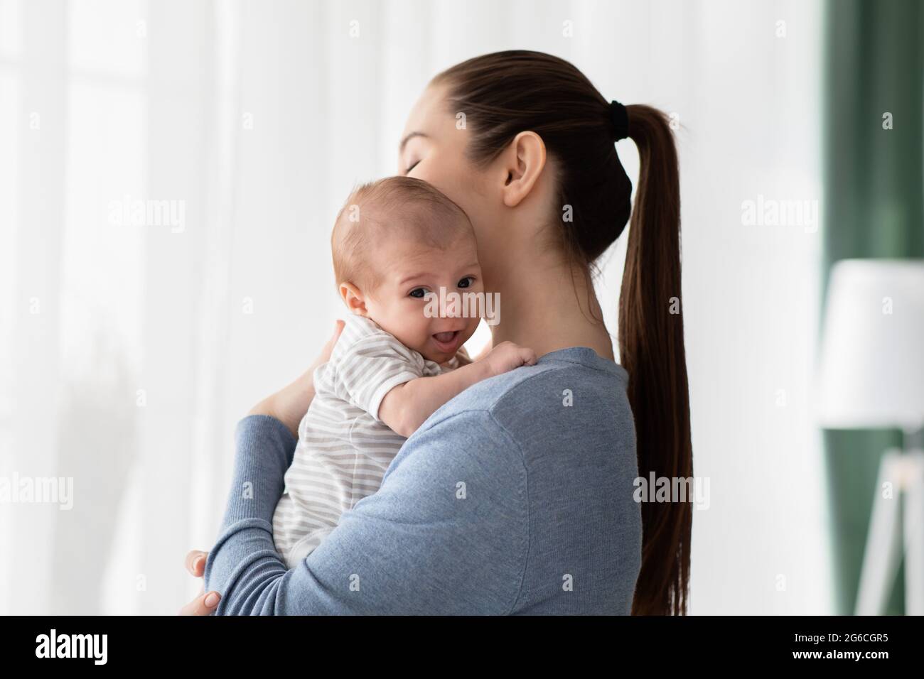 Young Mom Embracing Her Adorable Infant Child, Standing Near Window At ...