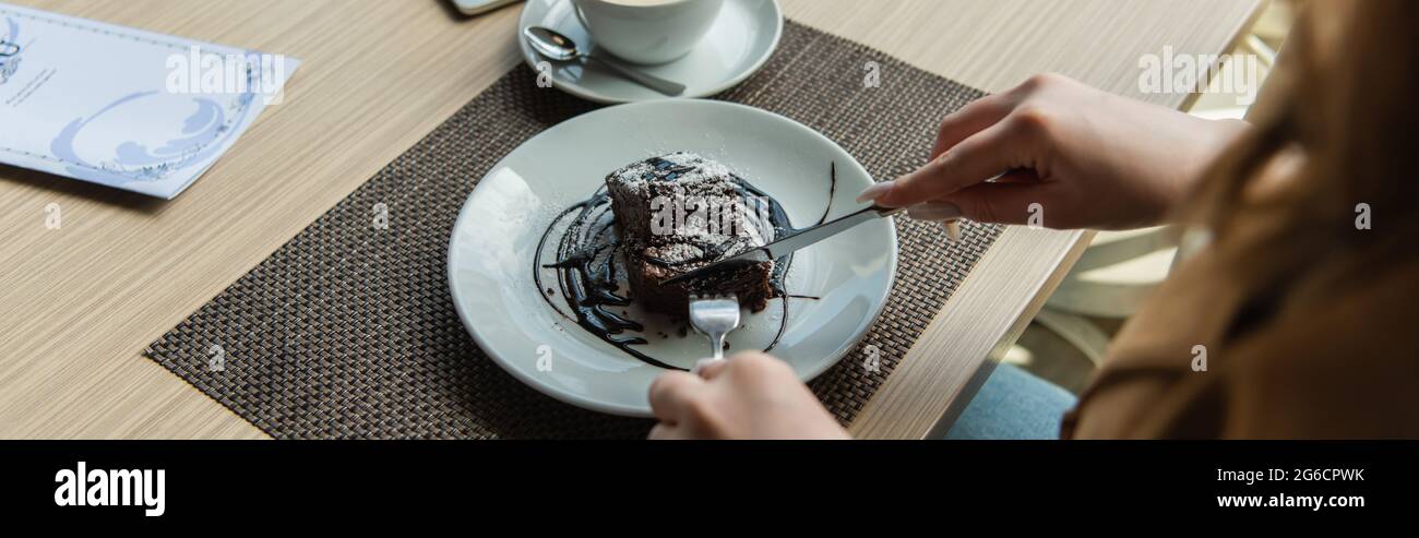 Cropped view of woman cutting dessert near cup and menu in cafe, banner ...
