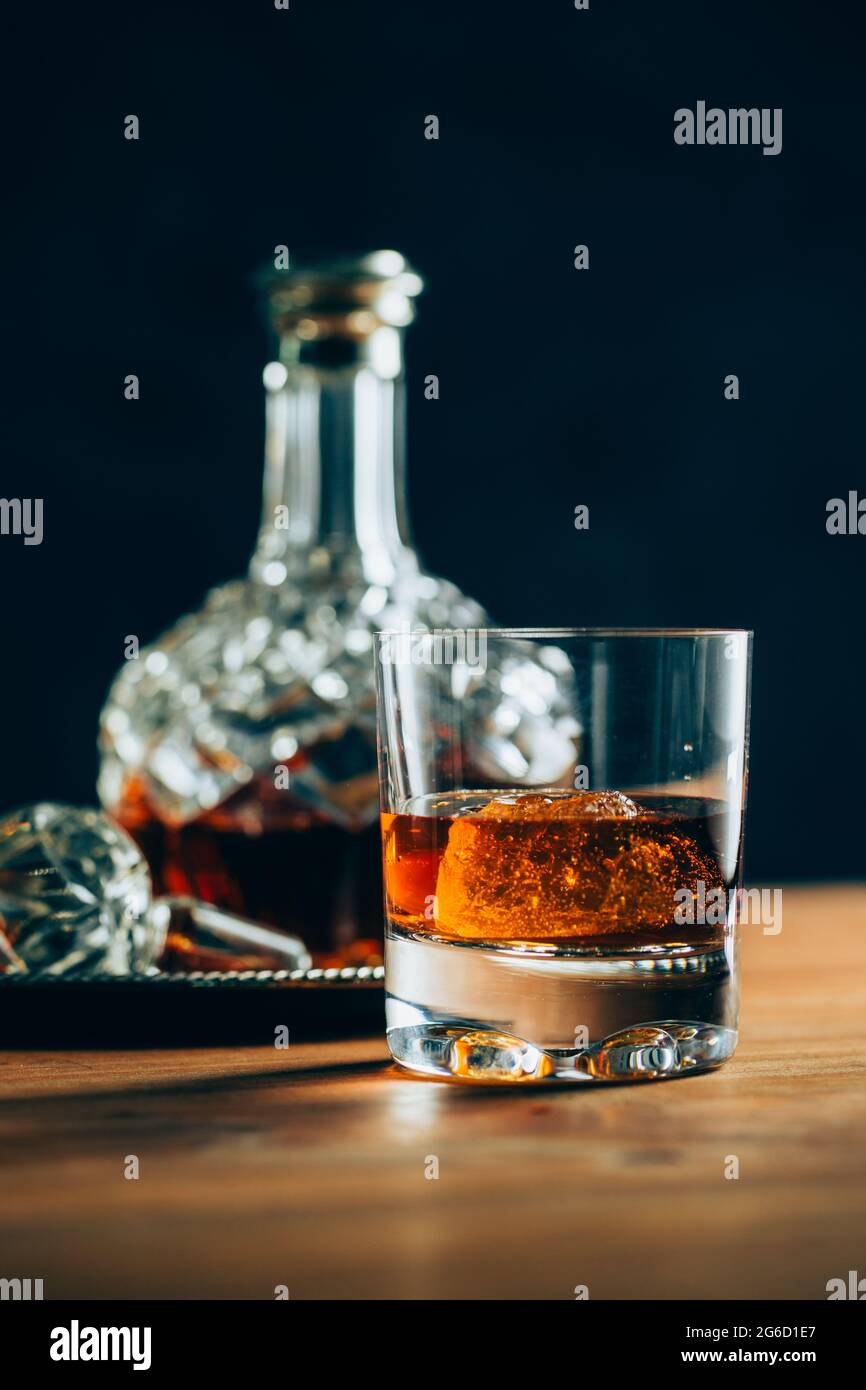 Glass Of Cold Whiskey With Ice Placed On Wooden Table Near Decanter On Black Background Stock