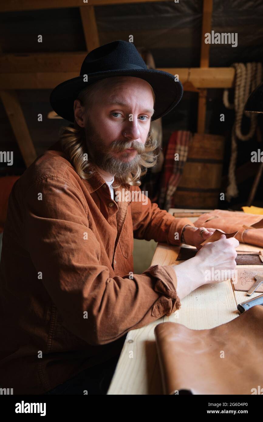 Portrait of shoemaker in hat looking at camera while repairing shoes at ...