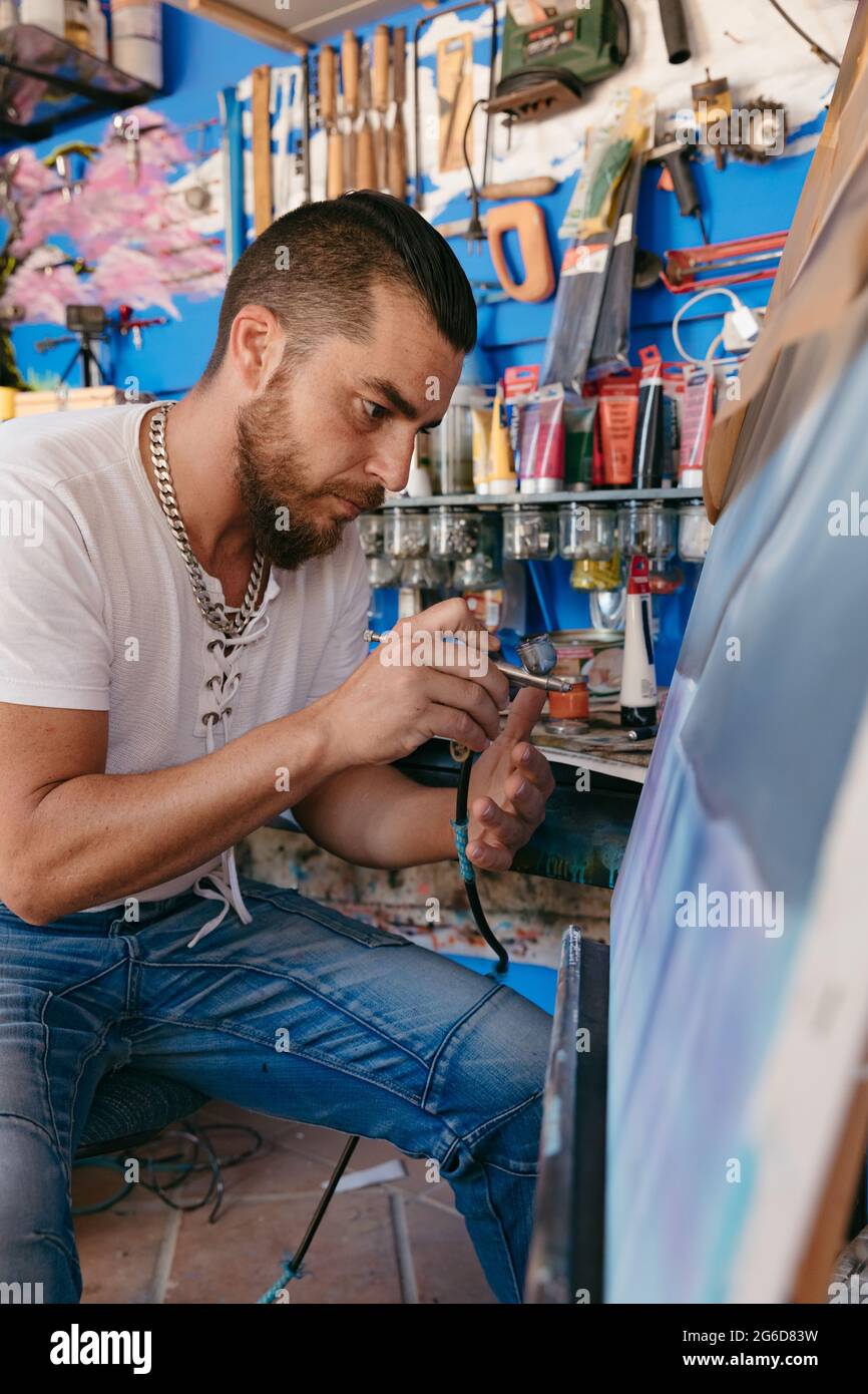 Side view of male artist using spray gun to paint picture on canvas ...