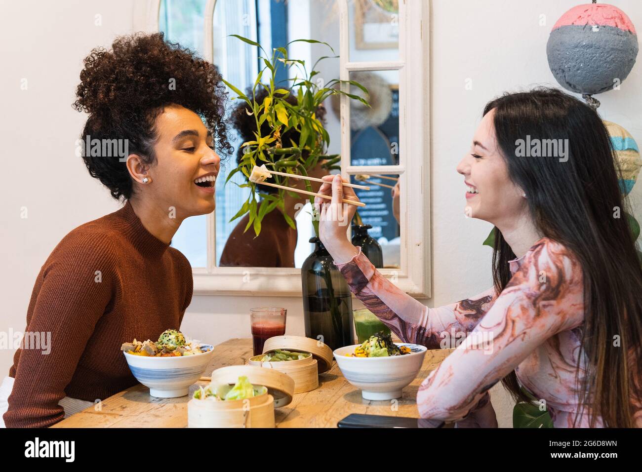 Side view of cheerful Woman feeding black female friend with poke while ...