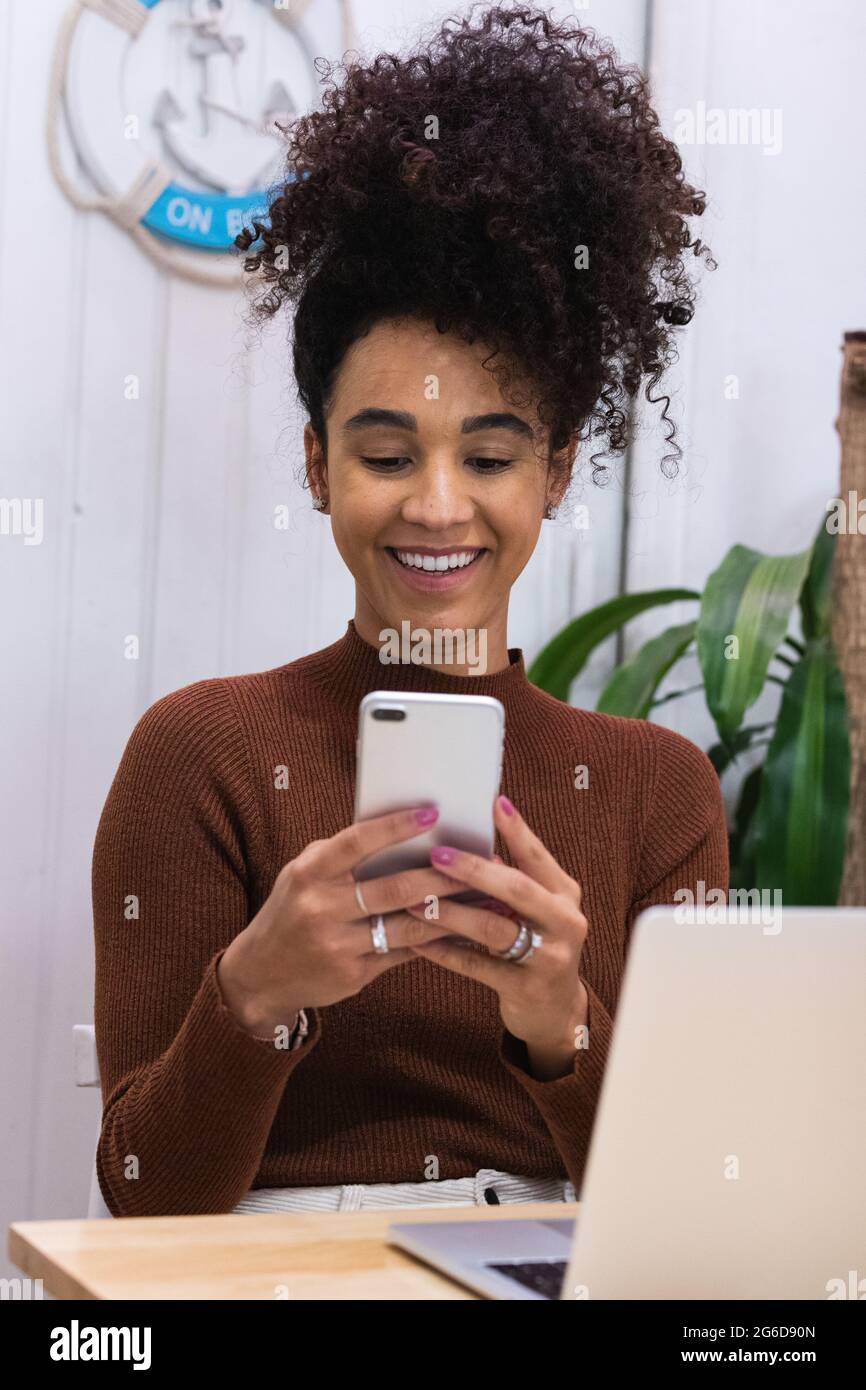 Happy African American female freelancer browsing smartphone and laughing while sitting at table ...