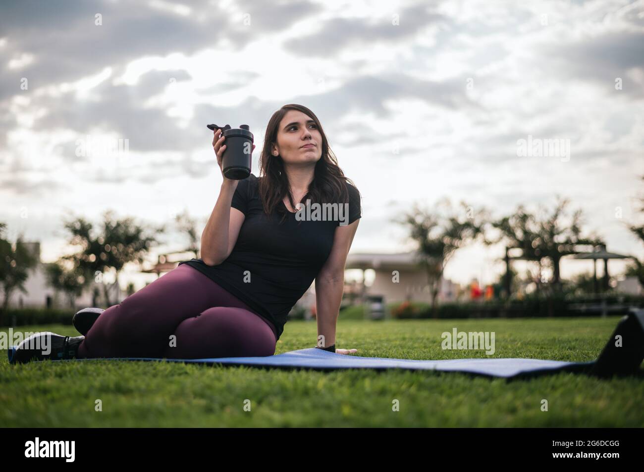 Ethnic athletic female sitting on mat on lawn and taking break during ...