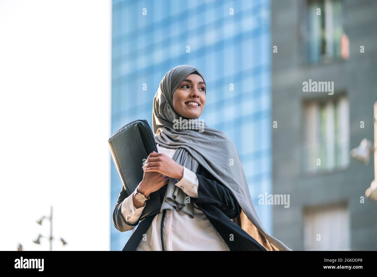 Cheerful Muslim female entrepreneur in hijab and with folder standing ...