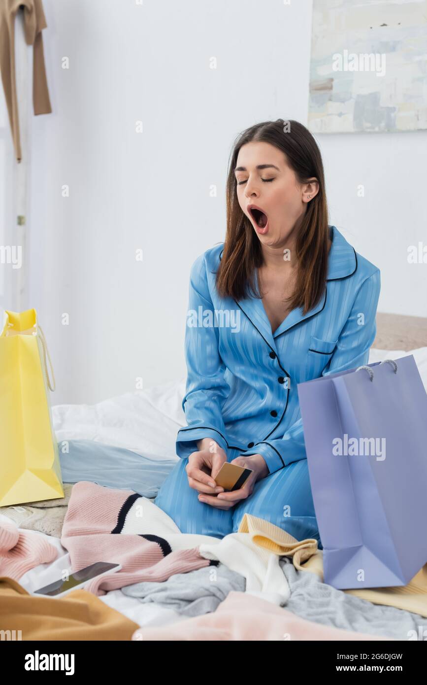 sleepy woman in stylish pajama yawning while sitting with credit card ...