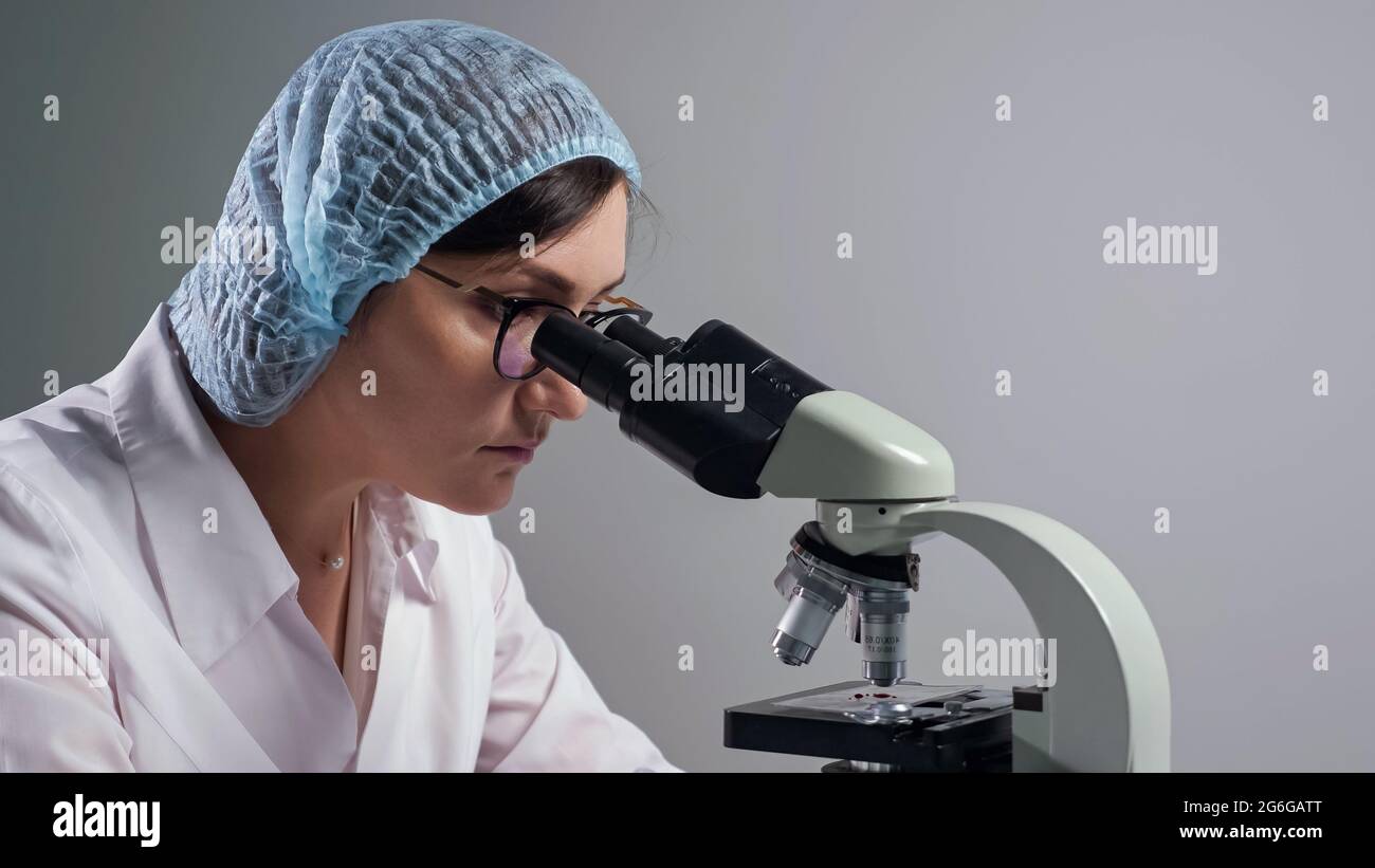 Female scientist with glasses in white coat and blue cap looks at ...