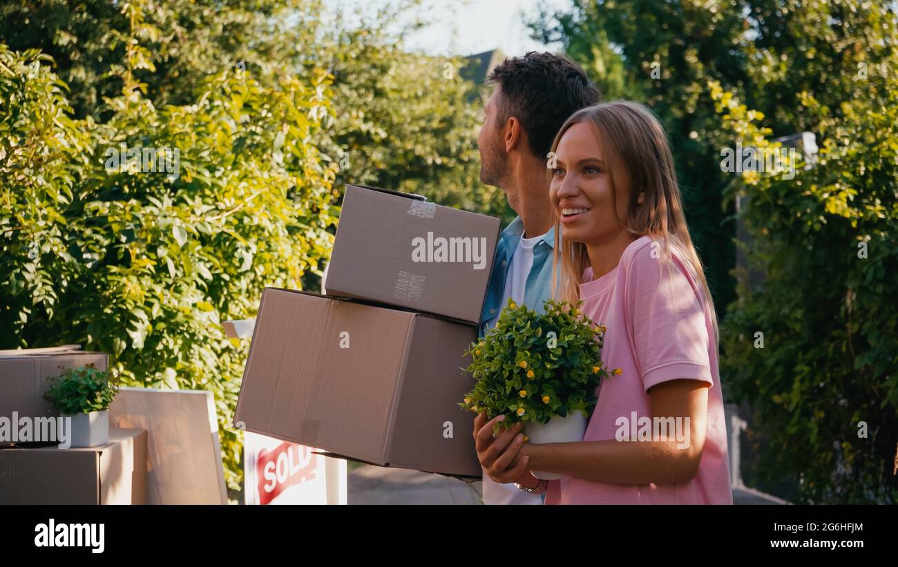 happy husband and wife walking with carton boxes and plant, relocation ...