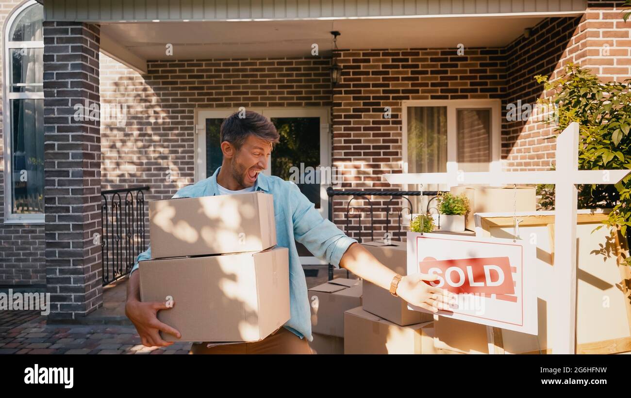 excited man holding boxes and pointing with hand at sold board near ...