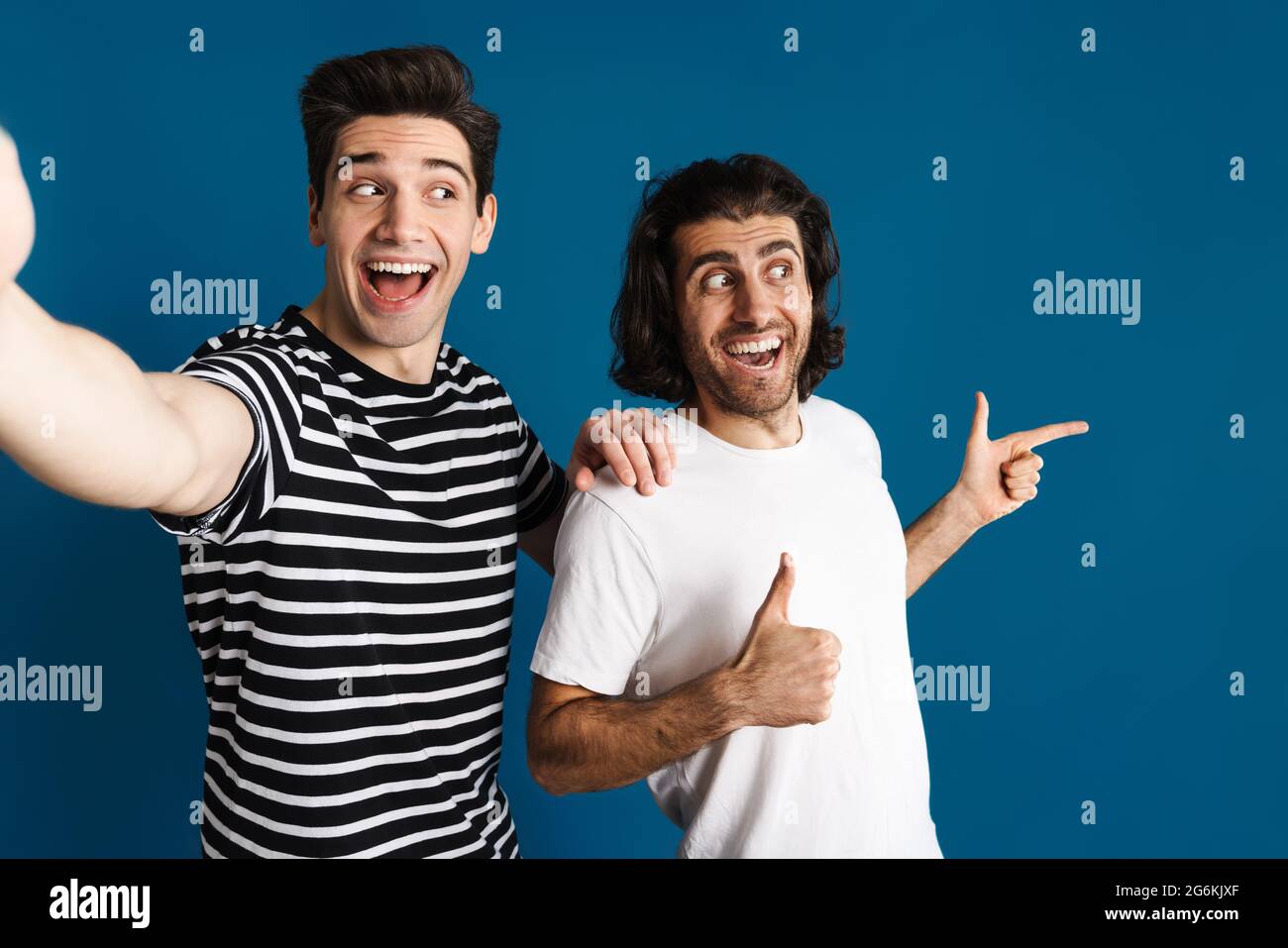 White excited two men gesturing thumb up while taking selfie photo isolated over blue background ...