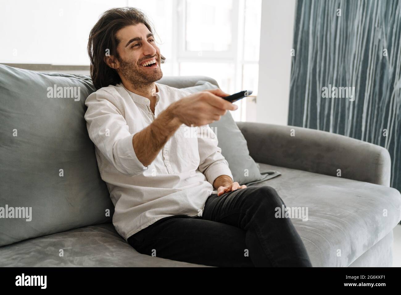 Happy young man holding TV remote while sitting on a couch at home and ...