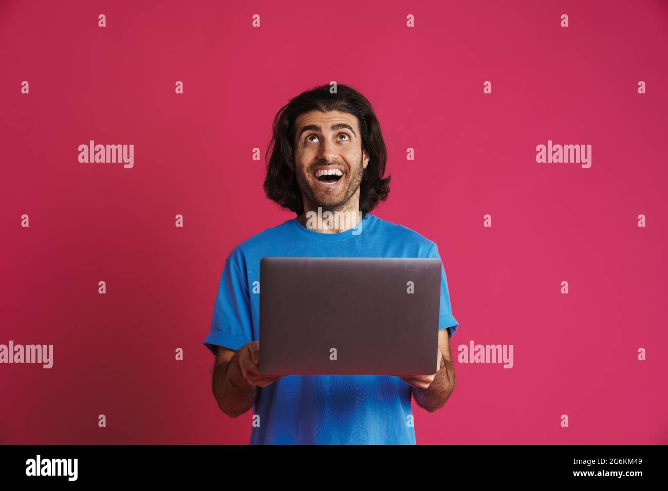 Unshaven excited man exclaiming and holding laptop isolated over pink ...