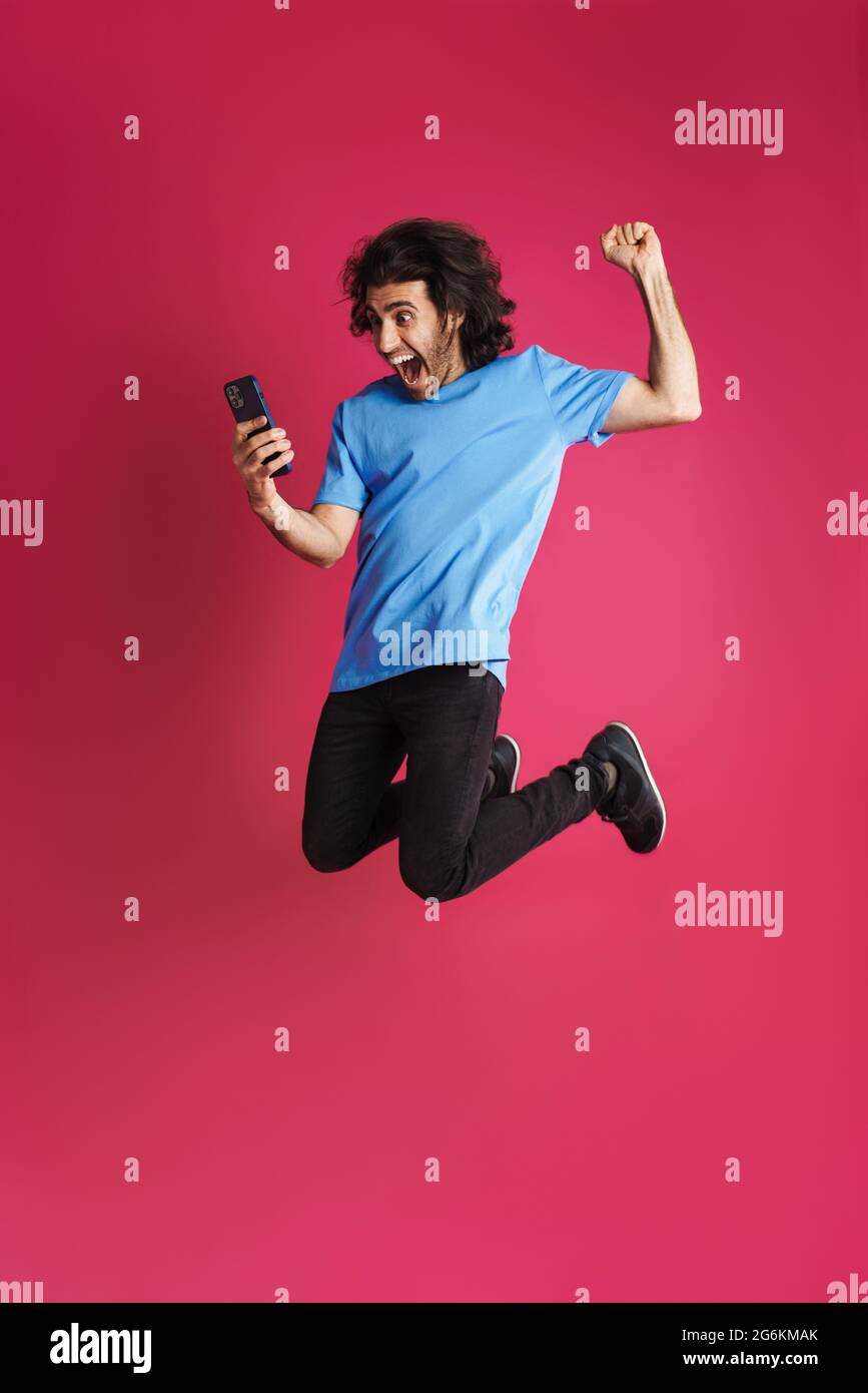 Excited brunette man jumping and using mobile phone isolated over pink ...