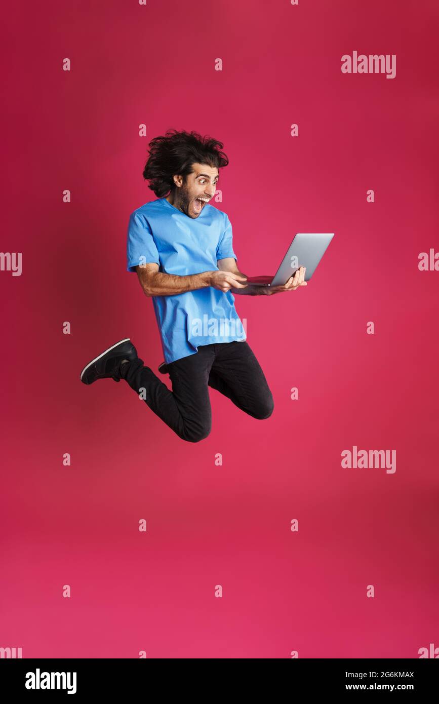 Excited brunette man screaming and using laptop while jumping isolated ...