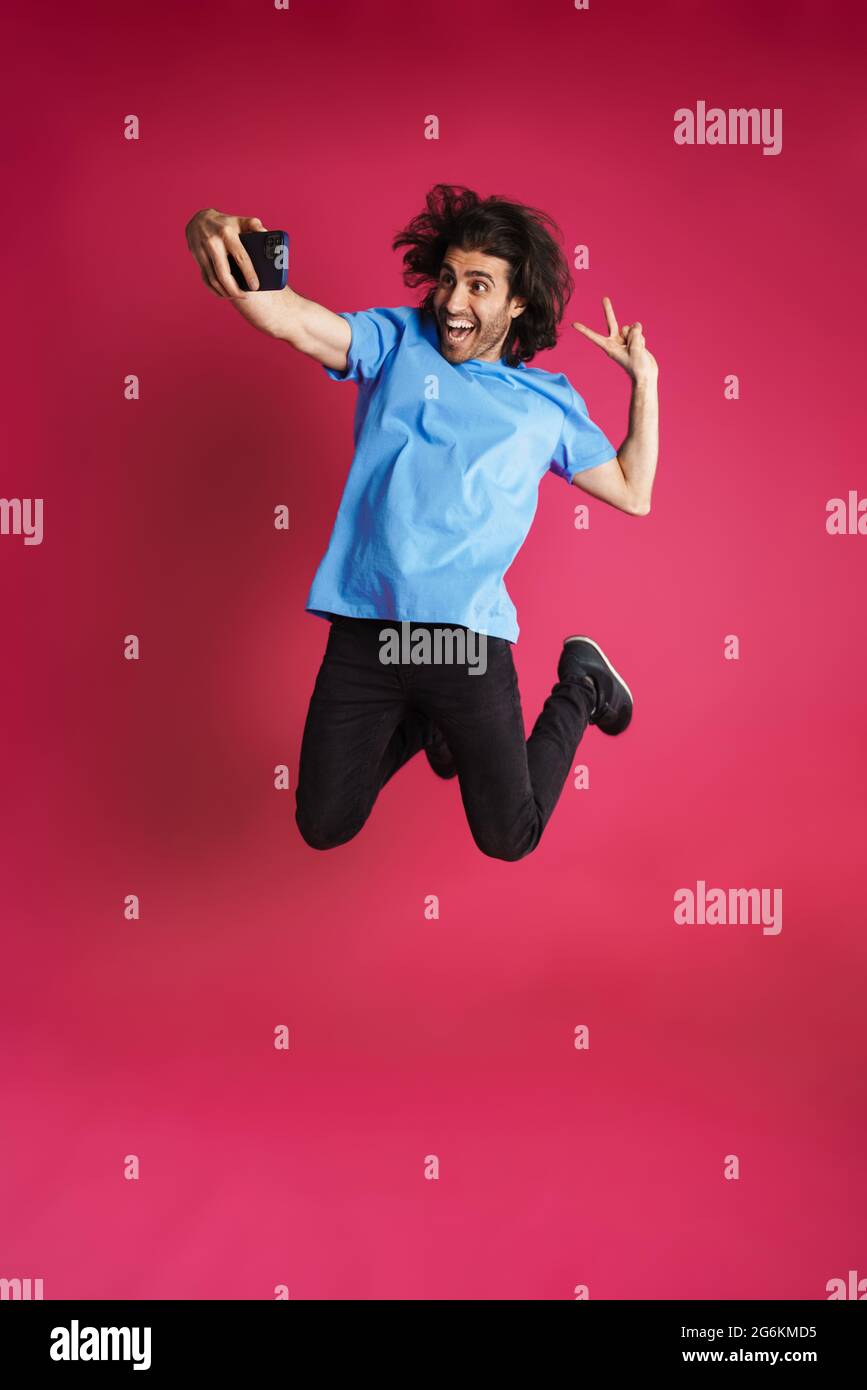 Excited brunette man jumping and using mobile phone isolated over pink ...